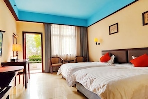 Quarto de hotel com duas camas, uma escrivaninha, cadeiras, um abajur e vista para o verde.