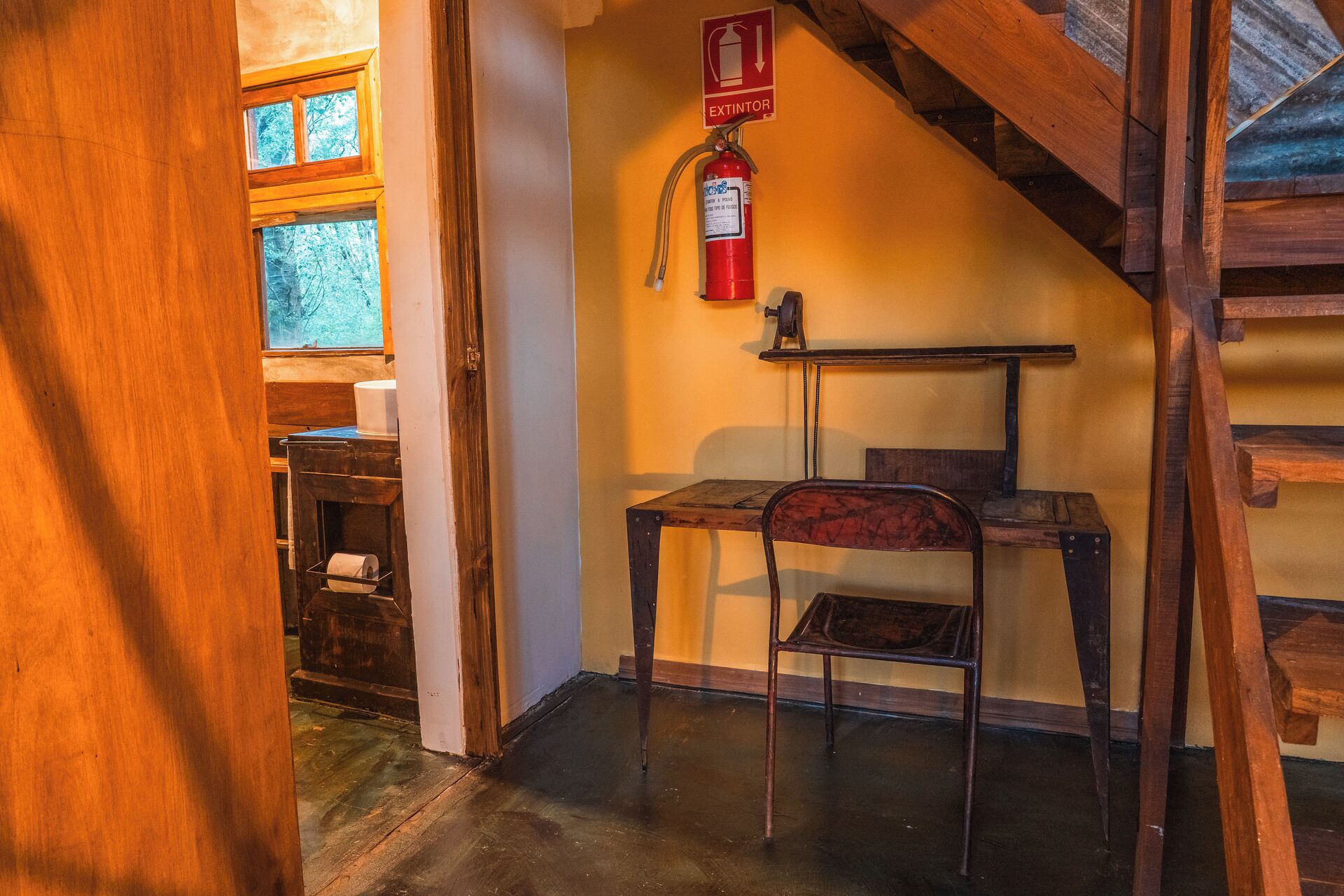 Um corredor estreito com uma mesa e uma cadeira de madeira, um extintor preso na parede e uma janela com vista para árvores.