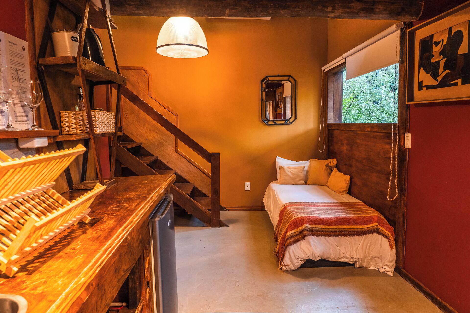 Um quarto aconchegante com uma escada de madeira, uma cama com um cobertor estampado, uma mesinha com um abajur e uma janela com vista para vegetação.
