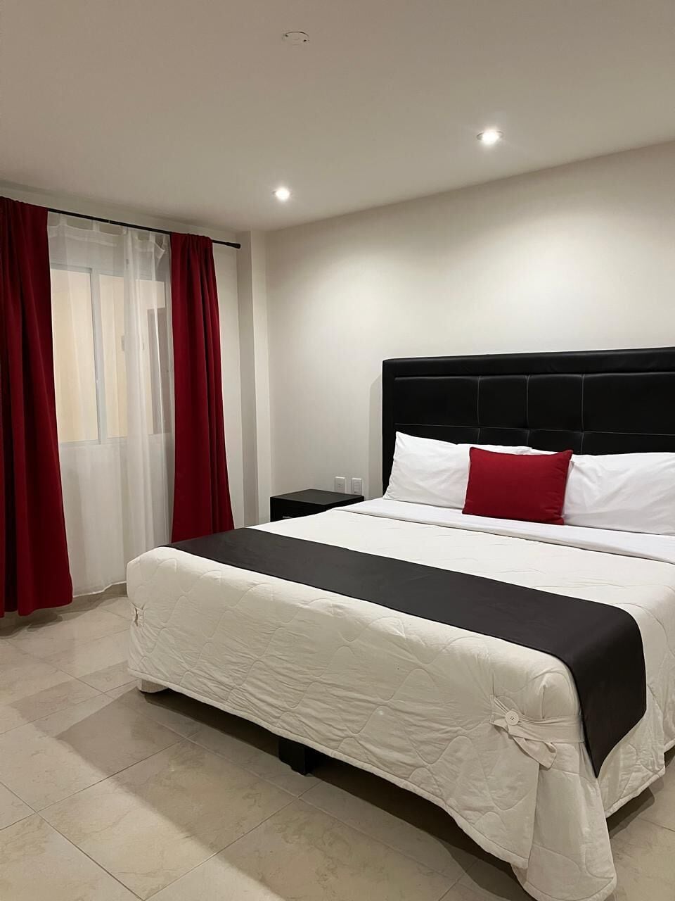 Quarto de hotel com uma cama grande, mesa de cabeceira e janela com cortinas vermelhas.