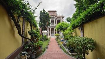 Um caminho leva a um edifício com varanda, cercado por vegetação e plantas em vasos.