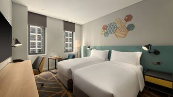 Quarto de hotel com duas camas, uma escrivaninha, uma cadeira e uma pequena mesa. Há janelas e um enfeite na parede.