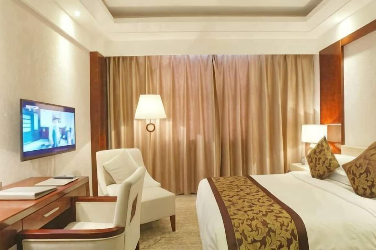 Quarto de hotel com uma cama, uma escrivaninha com cadeira, uma televisão e um abajur.