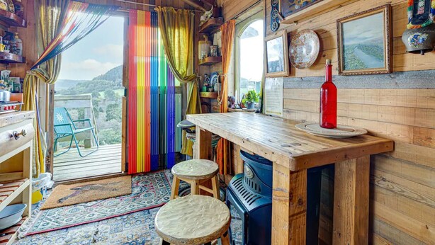 Interior rústico de uma cabana, com um balcão de madeira, dois banquinhos, uma garrafa vermelha e vista para as montanhas pela janela.