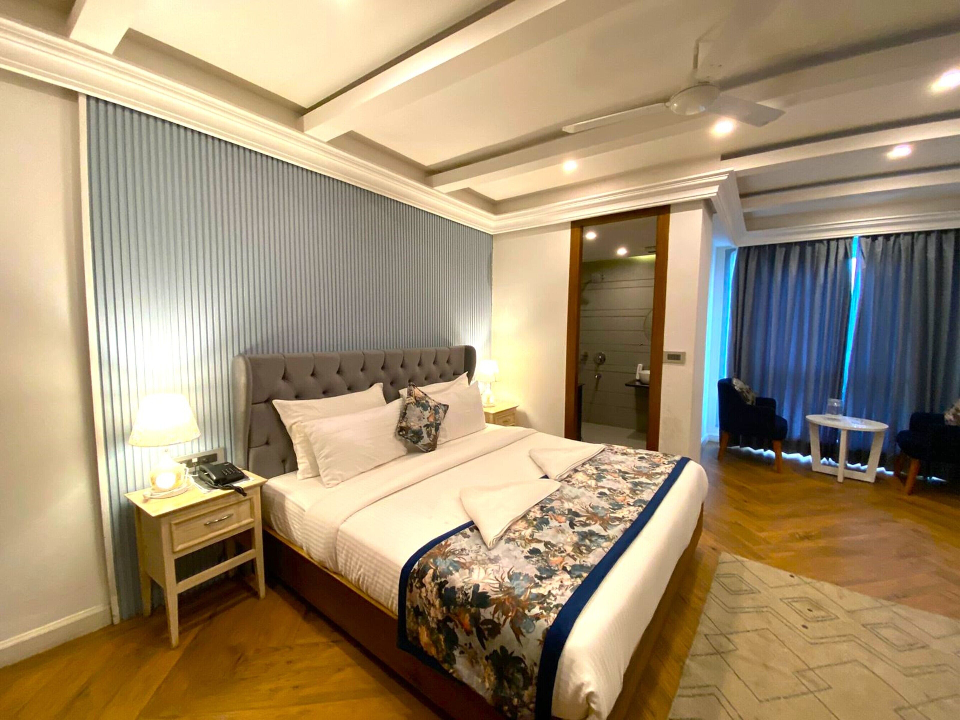 Quarto de hotel com uma cama grande, uma mesa de cabeceira com abajur, piso de madeira e vista para o banheiro através de uma porta aberta.