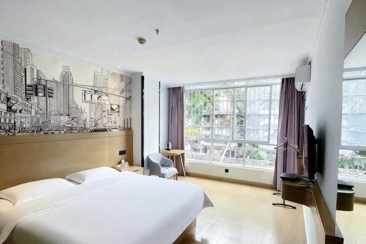 Quarto de hotel com uma cama grande, uma escrivaninha, uma cadeira, uma televisão e uma janela com vista para edifícios.