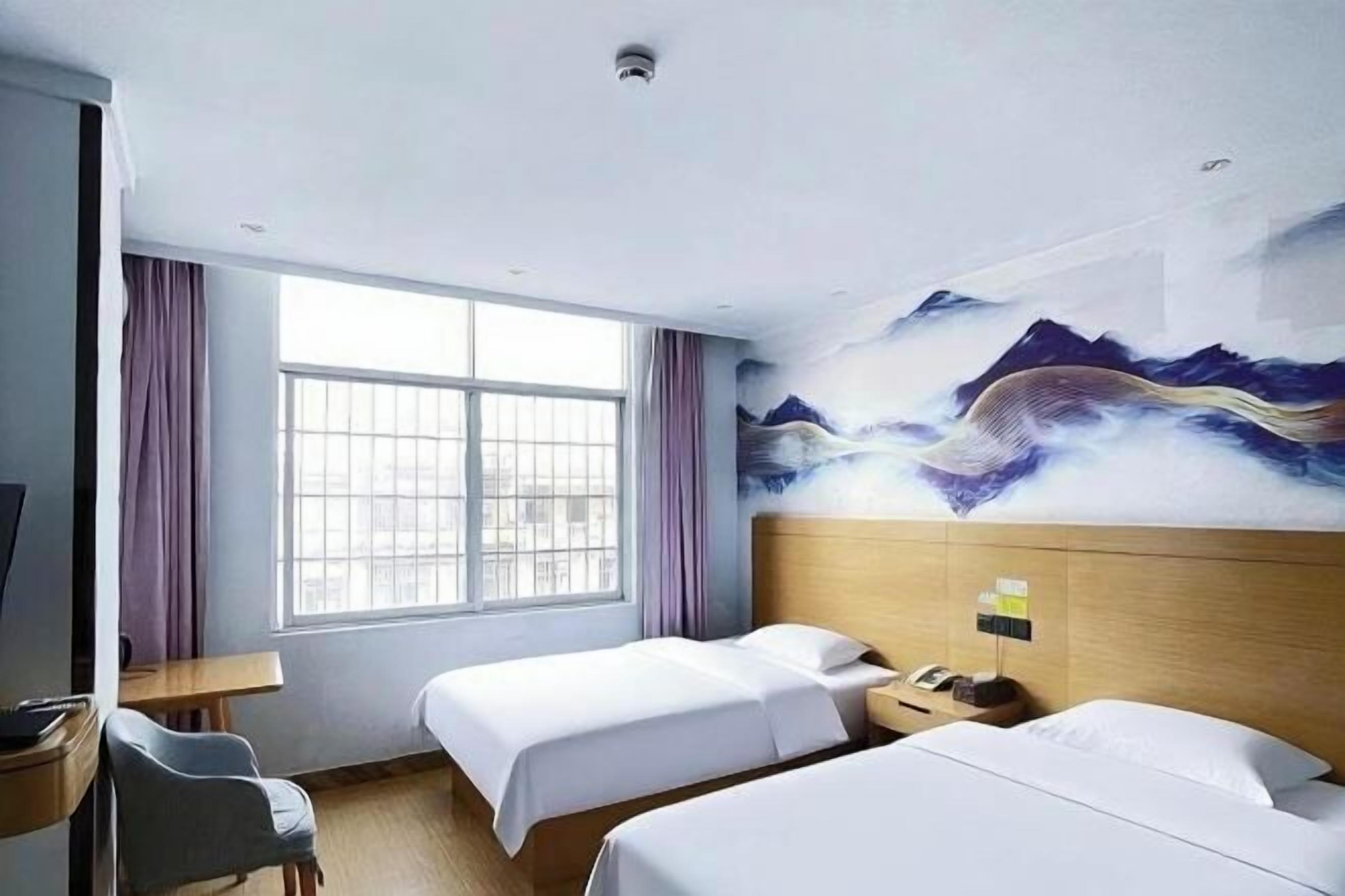 Quarto de hotel com duas camas, uma escrivaninha, uma cadeira, uma televisão, uma janela com cortinas e um mural na parede.