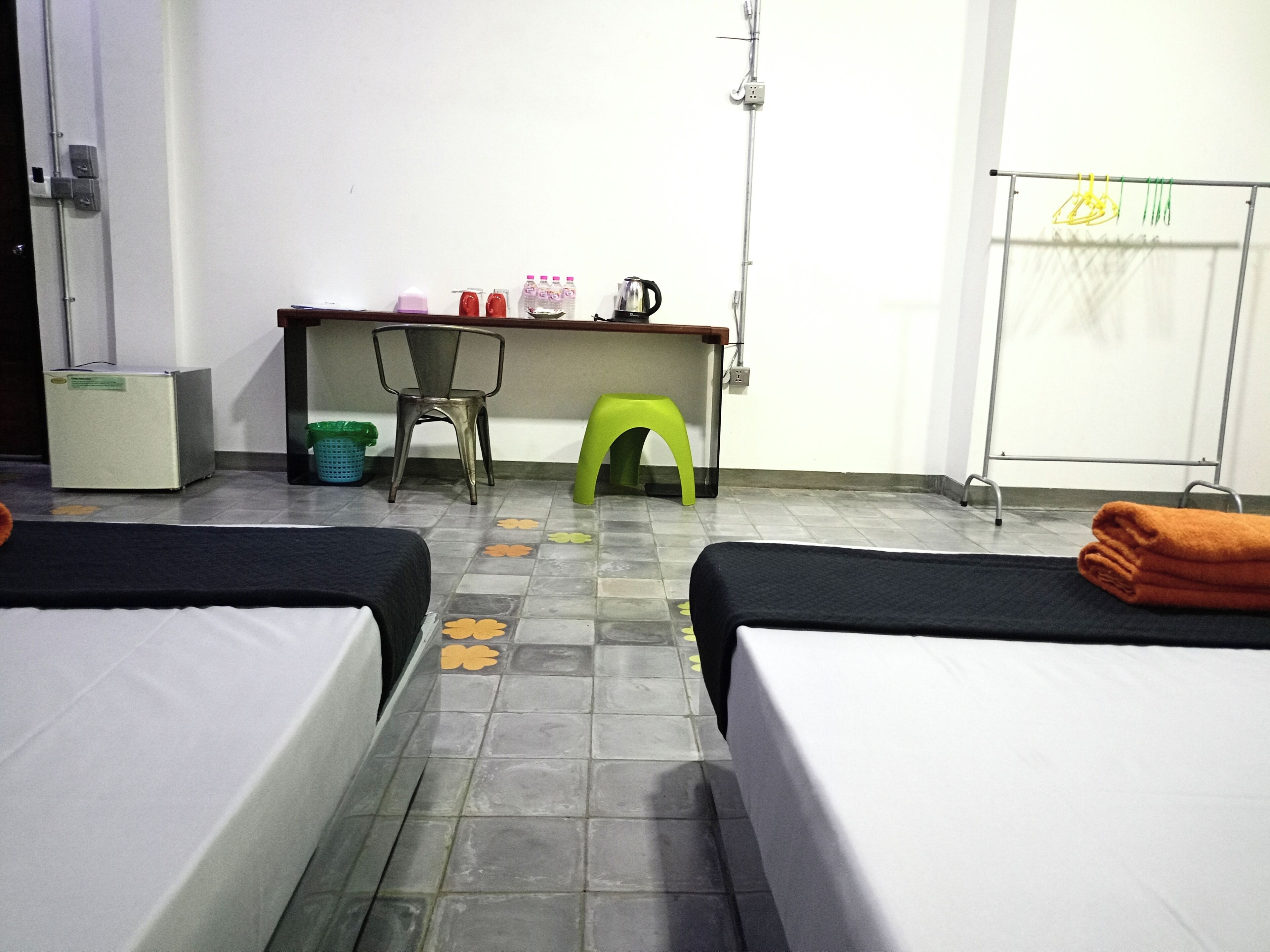 Quarto de hotel com duas camas, uma escrivaninha, uma cadeira, um mini-frigobar, um suporte para roupas e piso ladrilhado.