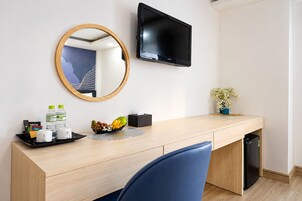 Quarto de hotel com escrivaninha de madeira, televisão na parede, espelho, uma pequena planta em vaso e um tabuleiro com artigos de higiene e uma fruteira.