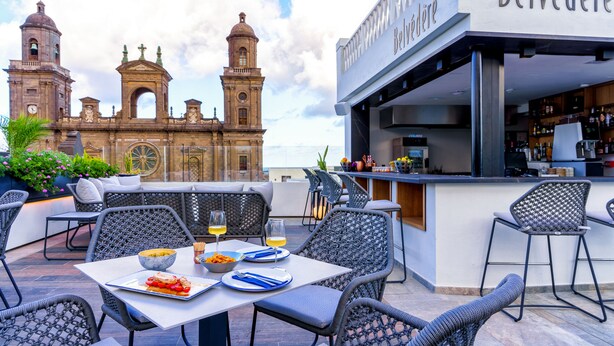 Uma área de refeições ao ar livre com cadeiras de vime e uma mesa posta com pratos e copos, um bar com uma placa indicando "Belvédère", e um edifício histórico com duas torres ao fundo.