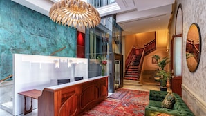 Um hall com uma mesa de recepção de madeira, um tapete com padrão vermelho, um sofá verde e uma escadaria para os andares superiores.