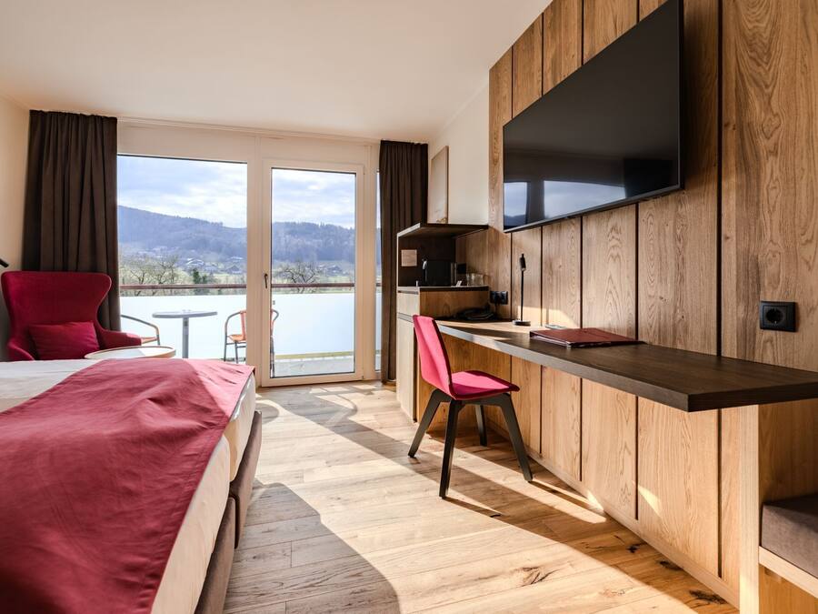 Quarto de hotel moderno com uma cama grande, uma escrivaninha com cadeira, televisão na parede e vista para um lago e montanhas.