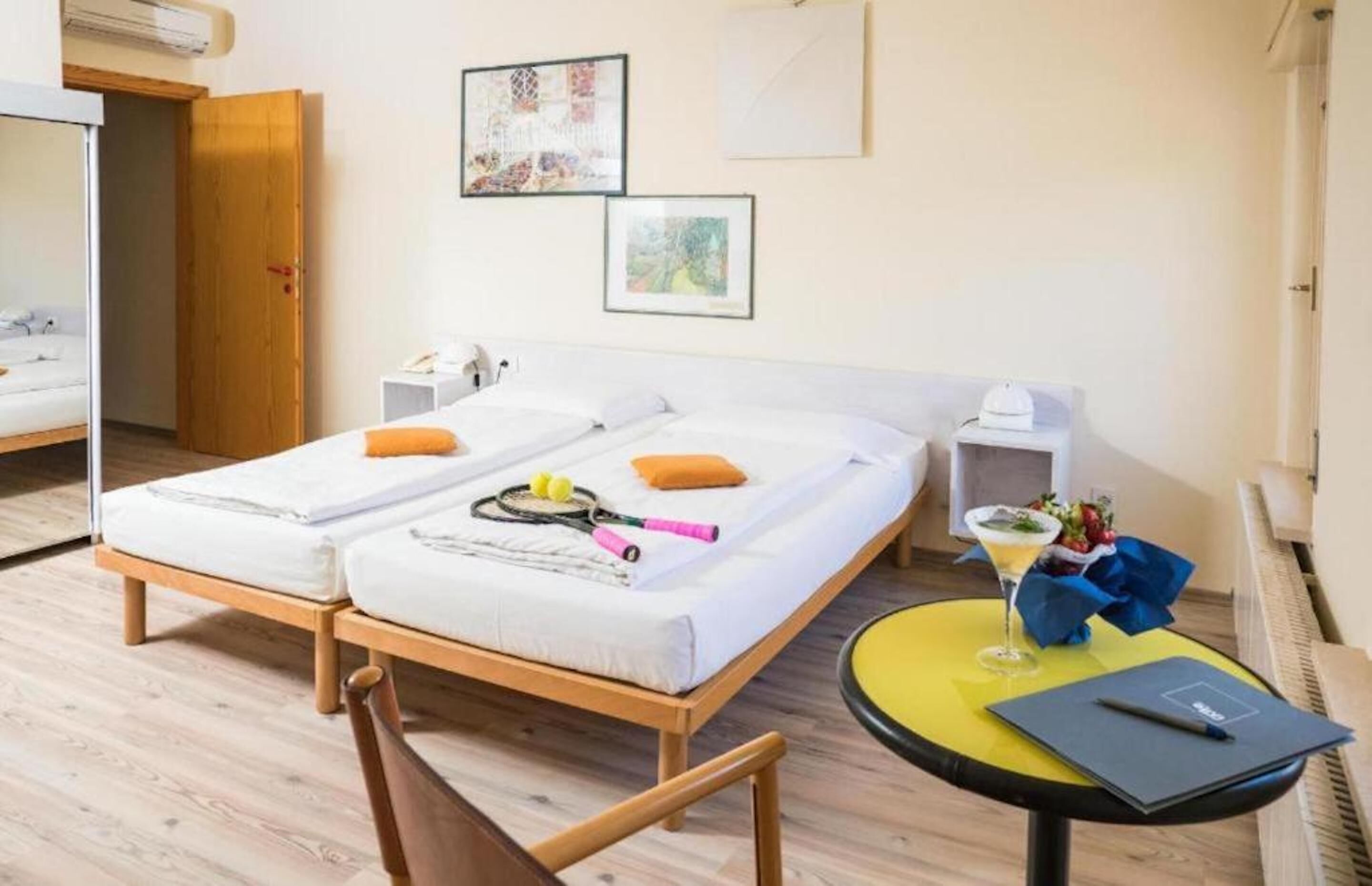 Quarto de hotel com duas camas, uma mesa pequena com uma bebida e um caderno, e obras de arte nas paredes.