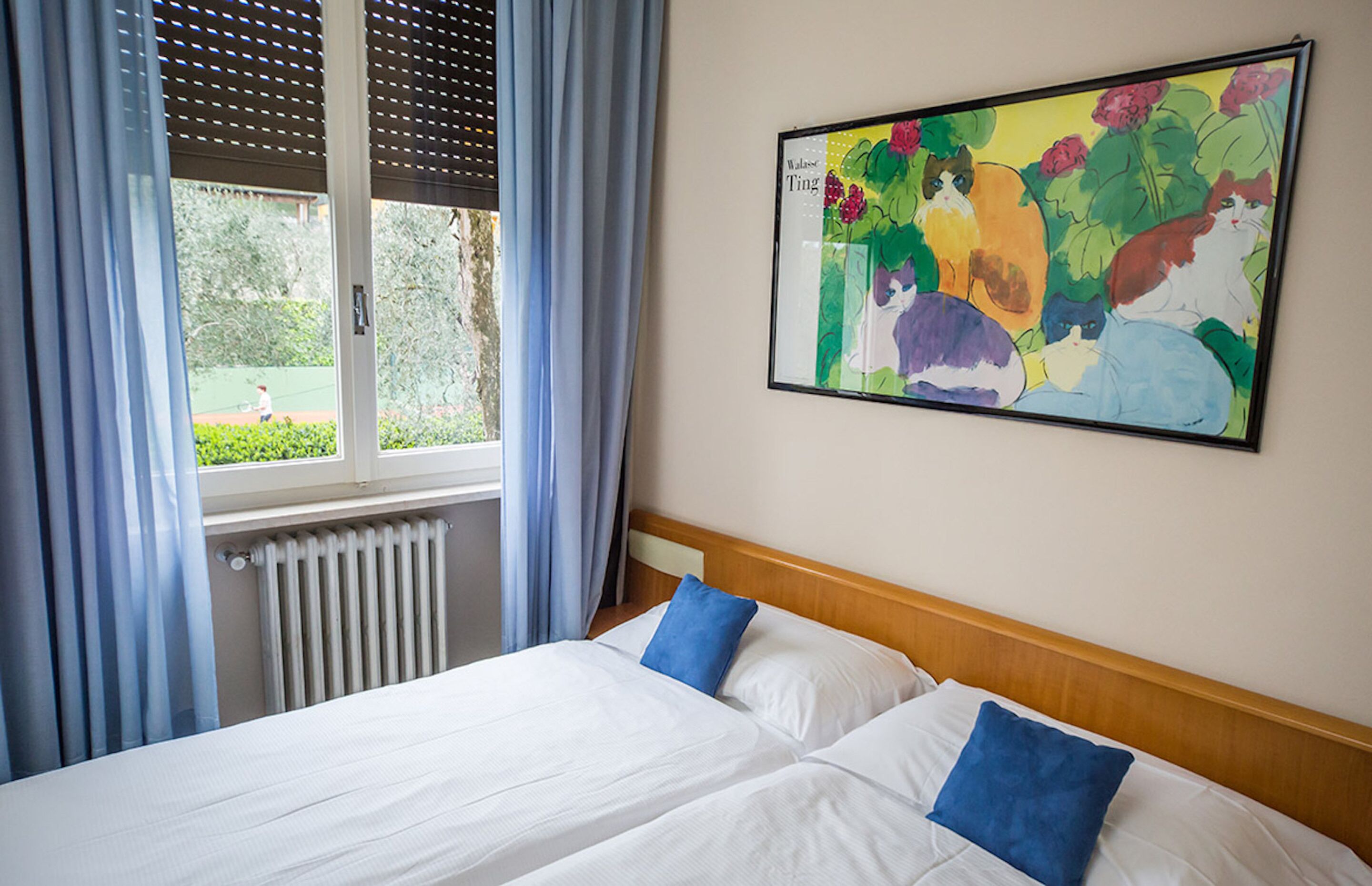Quarto de hotel com cama, duas almofadas, janela com cortinas e um quadro colorido na parede.