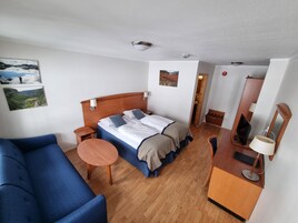 Quarto de hotel com uma cama, um sofá azul, uma mesinha redonda, uma escrivaninha de madeira, uma televisão e um espelho.
