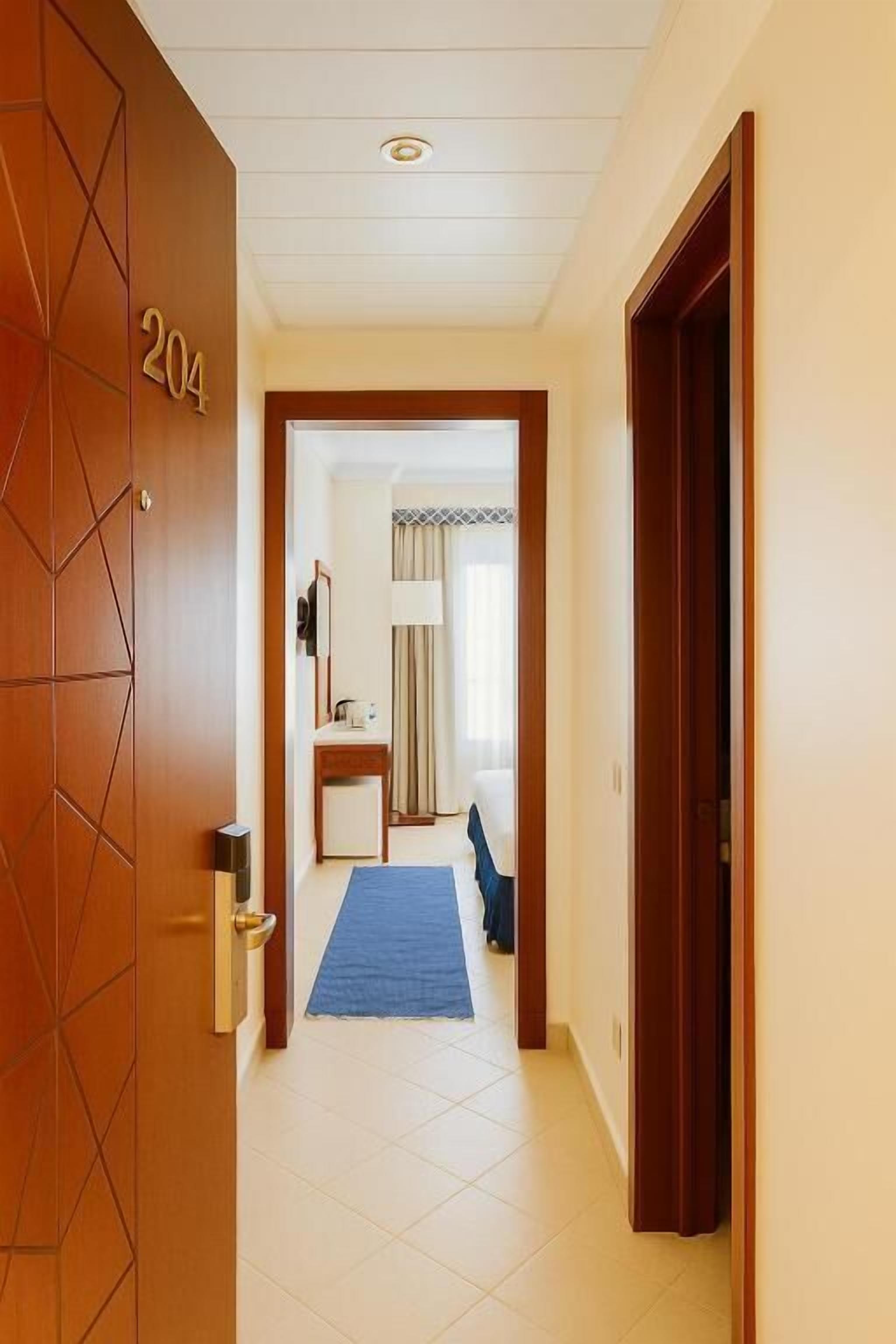 Corredor de hotel com uma porta de madeira identificada com o número 204, uma mesa com uma luminária, uma cama e um tapete azul.