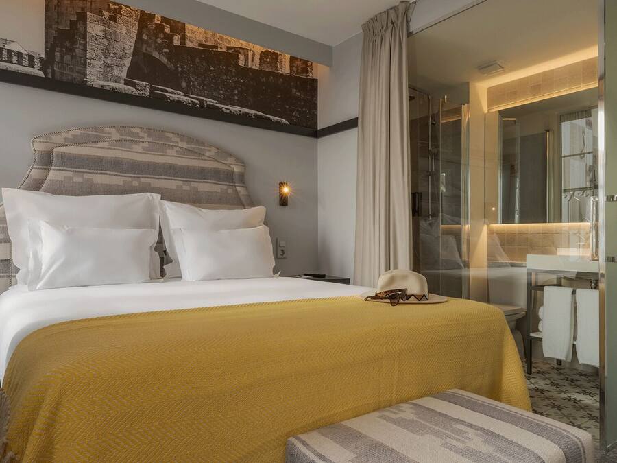 Quarto de hotel com uma cama grande, cabeceira com design texturizado, mesinha de cabeceira pequena, espelho e banheiro com pia e chuveiro.