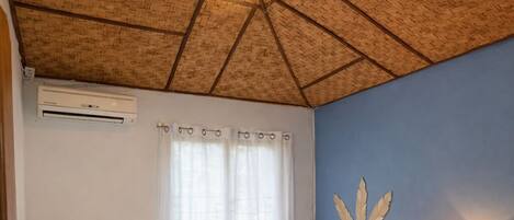 Quarto com teto de palha, cama, mesa de cabeceira com abajur, planta em vaso e ar condicionado.