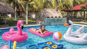 Uma piscina com brinquedos infláveis, palmeiras e uma cascata ao fundo.