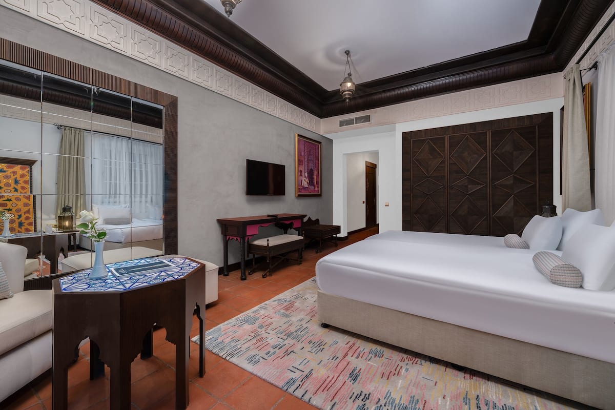 Quarto de hotel com cama, escrivaninha, cadeira, televisão e um espelho grande.