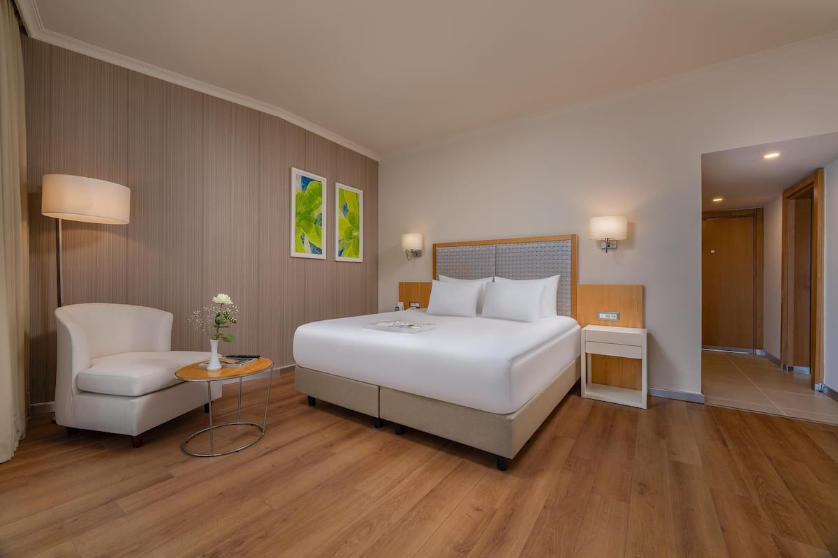 Quarto de hotel com uma cama grande, uma cadeira, uma mesa pequena com um vaso e duas luminárias na parede.