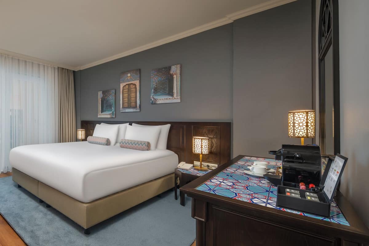 Quarto de hotel com cama, sofá, mesa pequena, escrivaninha com computador e televisão.