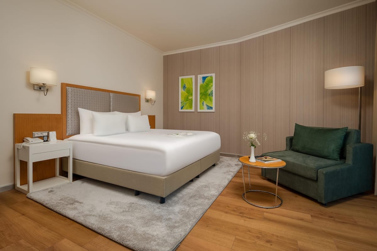 Quarto de hotel com uma cama, uma mesa de cabeceira, um sofá verde e duas luminárias fixadas na parede.