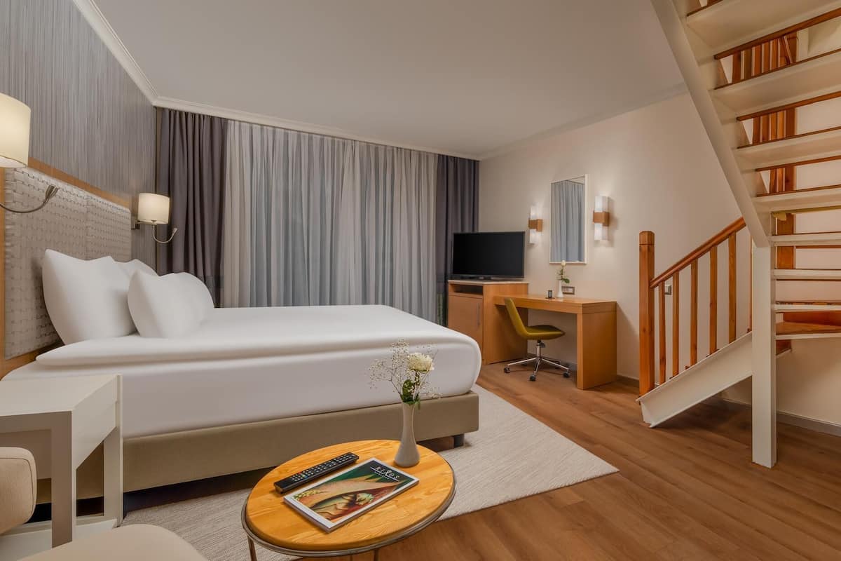 Quarto de hotel moderno com cama, mesa de cabeceira, televisão, escrivaninha, cadeira e escada.