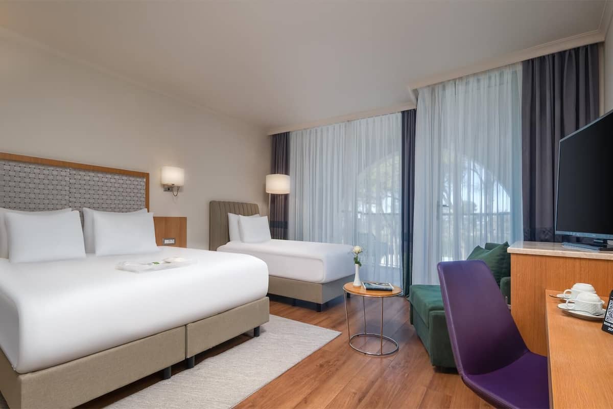 Quarto de hotel com uma cama grande, uma cama menor, uma mesa com cadeira, televisão e janela com cortinas.
