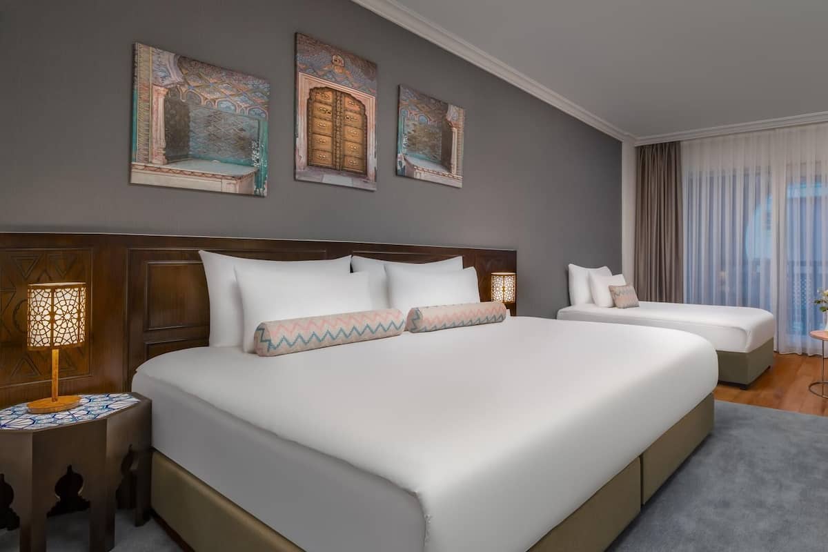 Quarto de hotel com uma cama grande, uma escrivaninha com chaleira, televisão e obras de arte nas paredes.