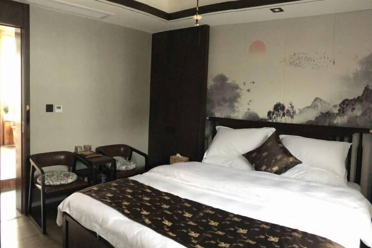 Quarto de hotel com cama, duas cadeiras, uma mesinha e parede com pintura.