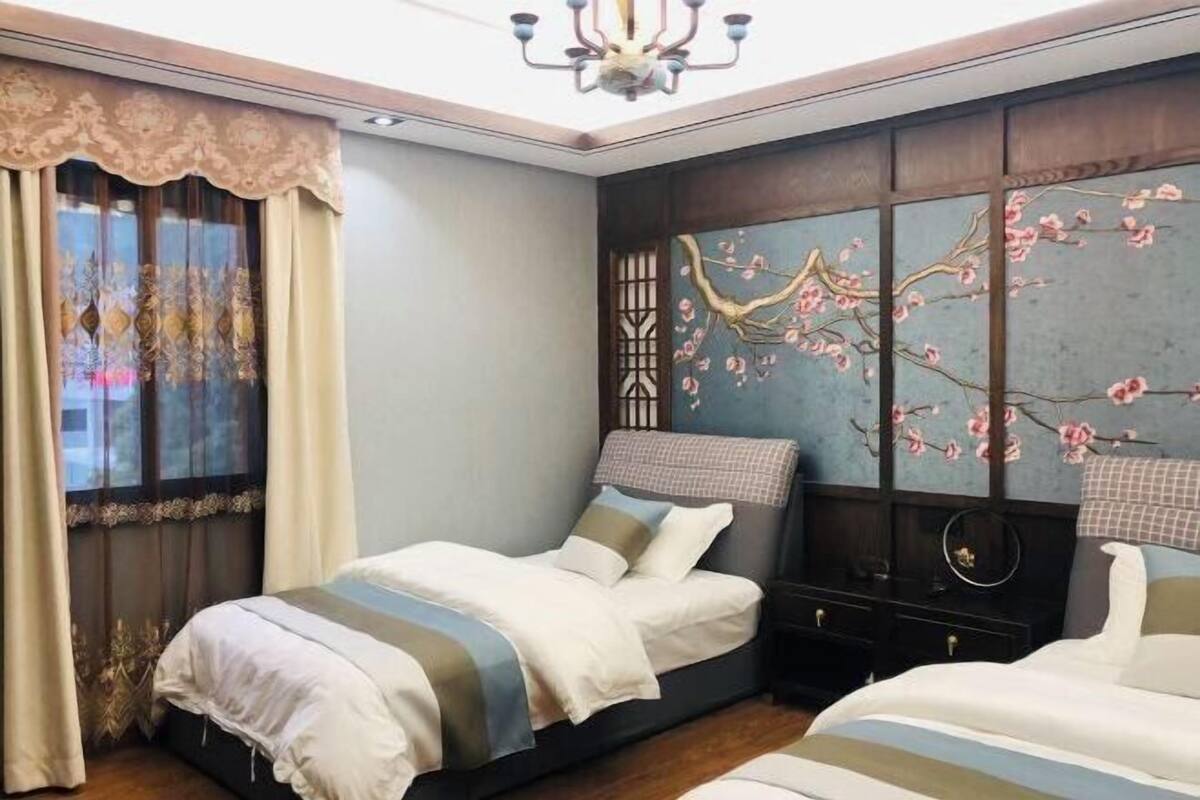 Quarto com duas camas, janela com cortinas, parede decorada com um mural floral e luminária no teto.