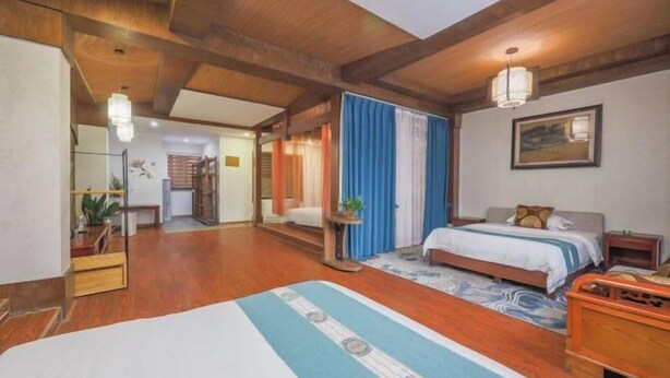 Quarto de hotel com teto de madeira, uma cama com colcha azul e branca, uma mesa de cabeceira e um abajur na parede, além de um quadro.