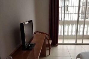 Quarto com escrivaninha de madeira, uma cadeira, televisão e vista para uma varanda com grade.