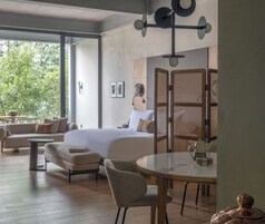 Quarto de hotel moderno com uma cama grande, área de estar com sofá e poltronas, mesa de jantar e vista para o verde lá fora.