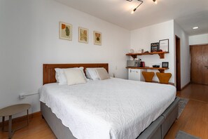 Quarto com uma cama grande, mesa de cabeceira, cadeira, prateleira com objetos e uma porta.