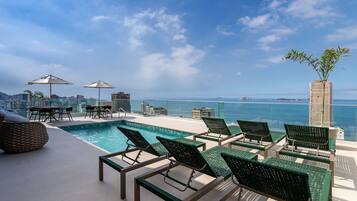Área de piscina no terraço com espreguiçadeiras e guarda-sóis, com vista para a paisagem urbana e o mar.