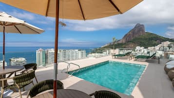 Piscina externa com espreguiçadeiras e guarda-sóis, com vista para uma paisagem urbana e uma montanha imponente.