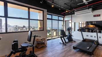 Academia com equipamentos de ginástica, amplas janelas e vista para a cidade.