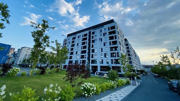 Um edifício residencial moderno, com várias andares e varandas, rodeado por vegetação e um céu claro.