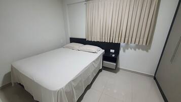 Quarto de hotel com cama, mesa de cabeceira, tomada e janela com cortinas.