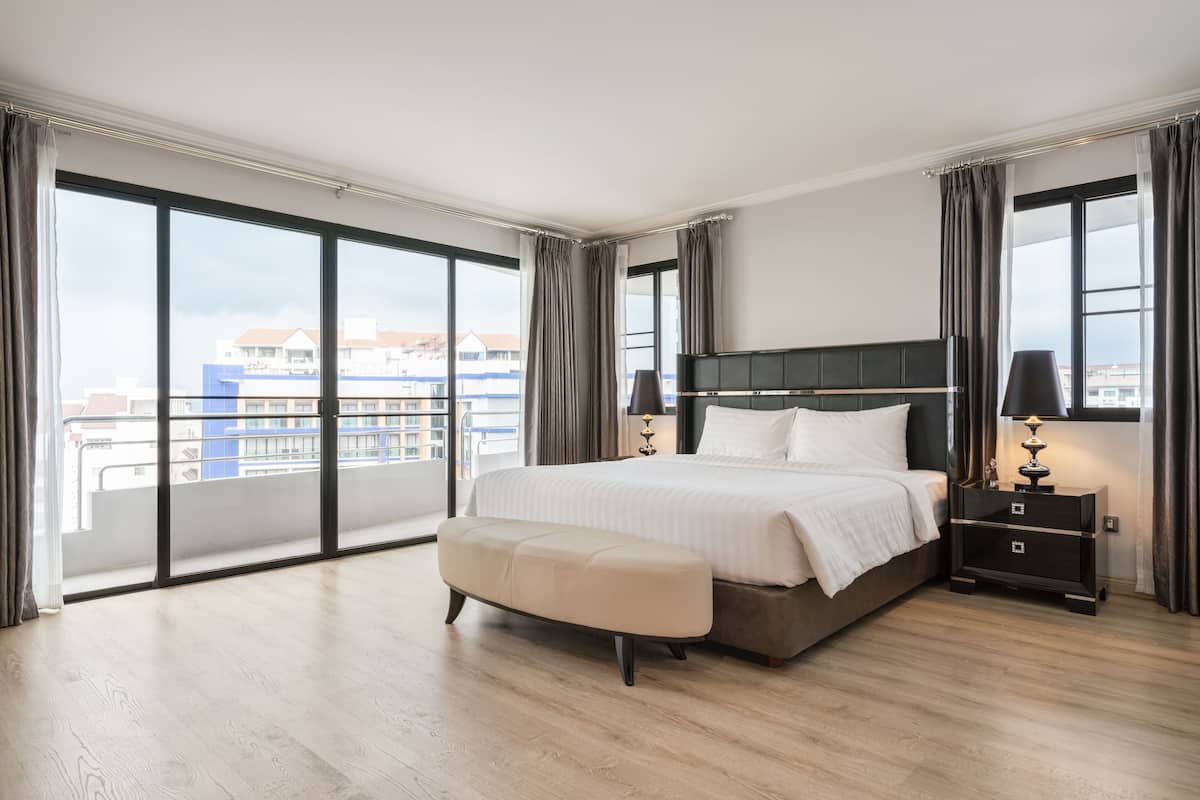 Quarto de hotel moderno com uma cama grande, um banco, uma mesa de cabeceira e vista da cidade através de janelas do chão ao teto.