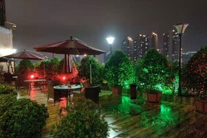 Área de terraço ao ar livre com mesas e guarda-sóis, rodeada por plantas em vasos e iluminada por iluminação ambiente.