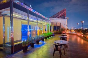 Uma área externa com mesas e cadeiras, plantas em vasos e um edifício com sinalização iluminada.