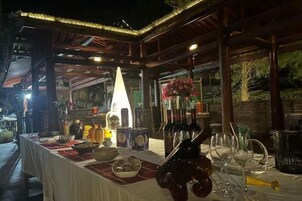 Uma área de refeições ao ar livre com uma mesa posta, cercada por vigas de madeira e uma parede de tijolos.