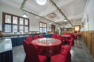 Uma sala de jantar com mesas redondas e cadeiras vermelhas, um buffet e grandes janelas que proporcionam luz natural.
