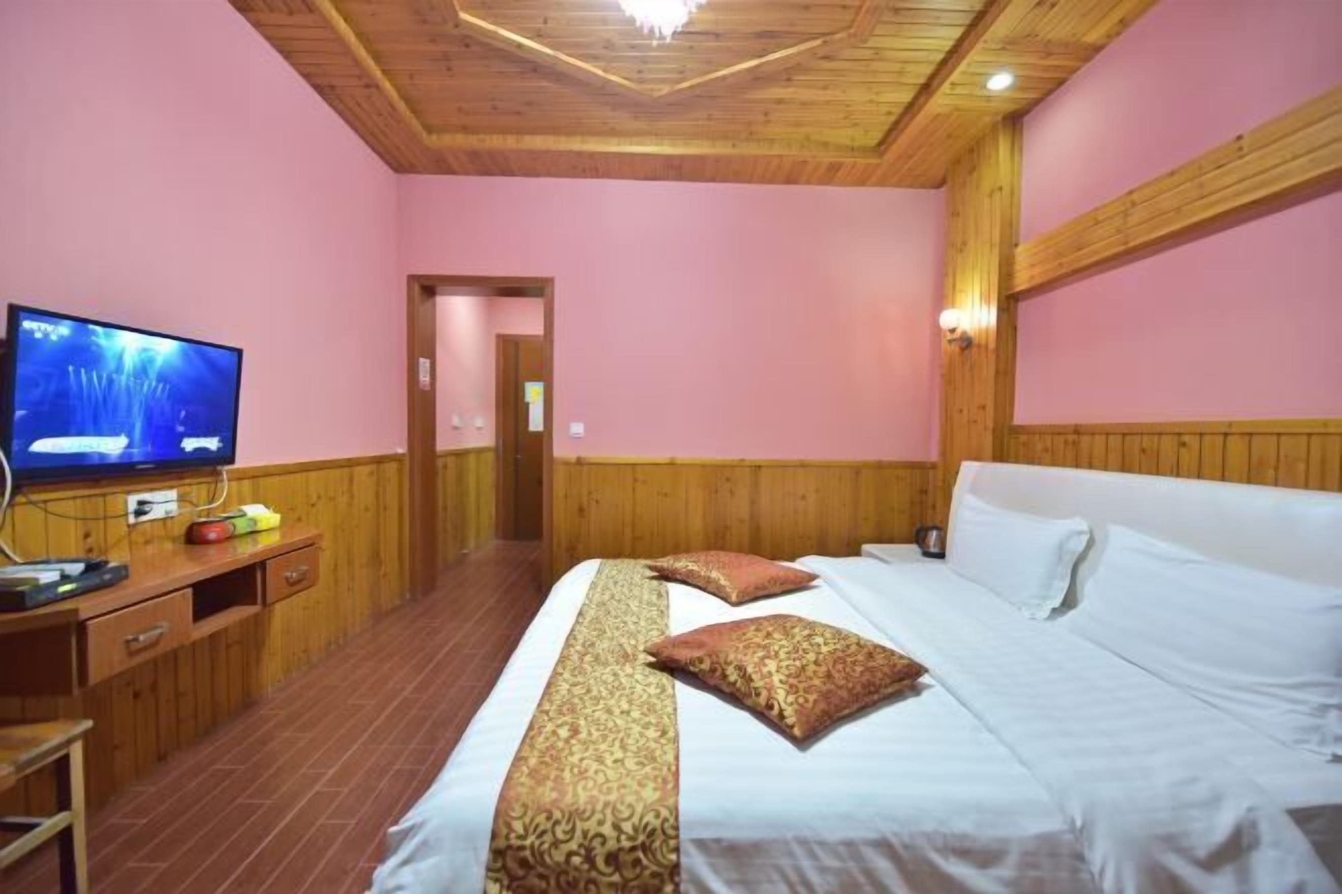 Quarto de hotel com teto de madeira, paredes cor-de-rosa, cama com roupa de cama branca, duas almofadas decorativas, uma TV em uma escrivaninha de madeira e uma mesa de cabeceira de madeira.