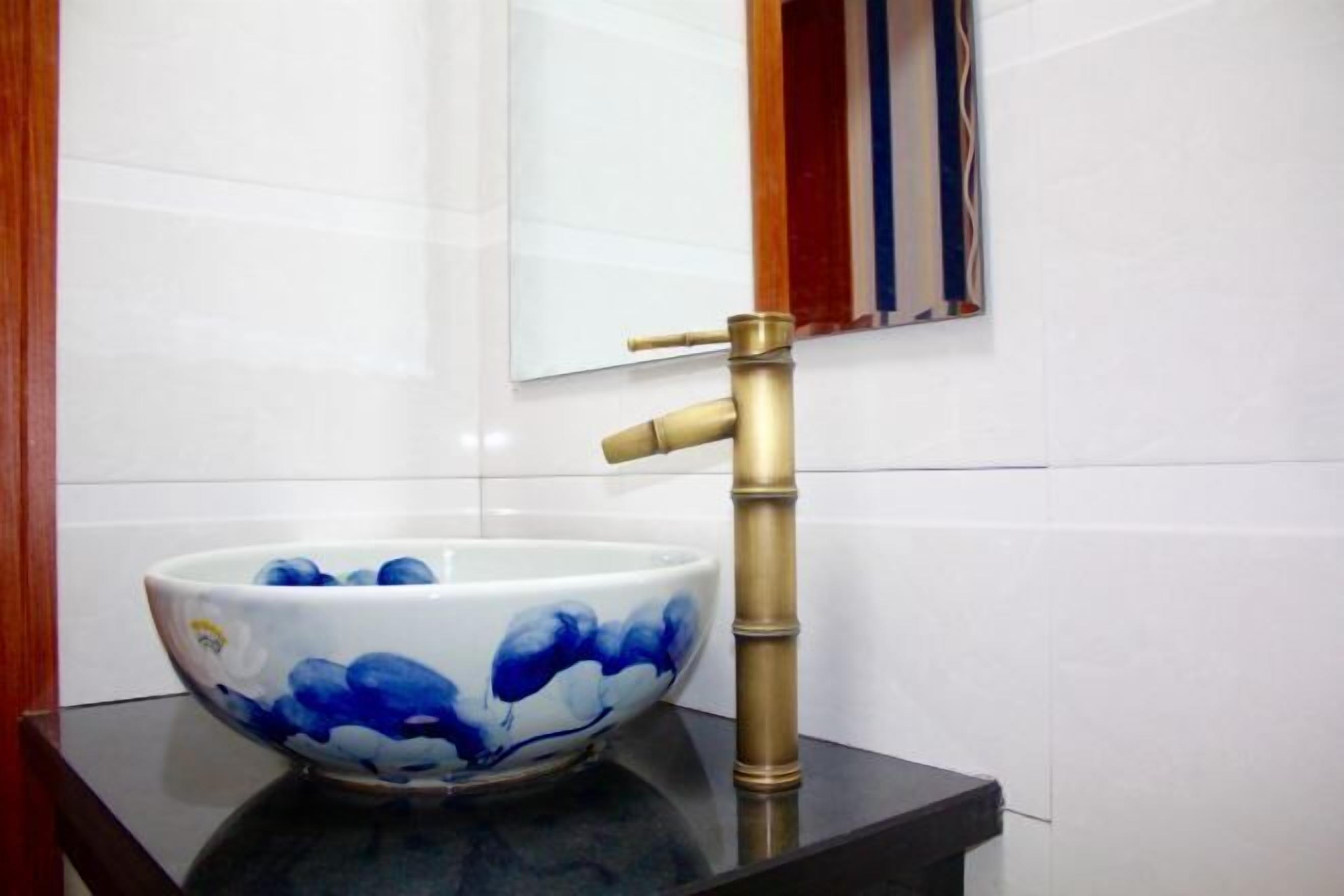 Casa de banho com parede de azulejos brancos, torneira dourada e uma tigela azul com motivos florais sobre bancada preta.