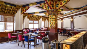 Um restaurante com decoração tradicional, incluindo pilares ornamentados e designs de teto estampados. As mesas estão dispostas para refeições, e há cadeiras com almofadas.