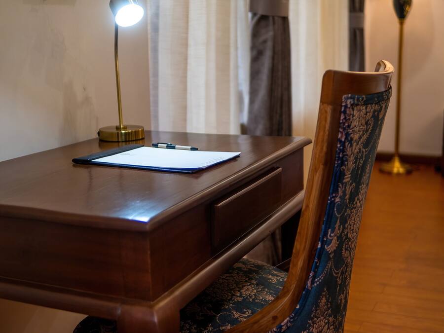Uma mesa de madeira com uma luminária, uma cadeira com almofada estampada e uma tomada na parede.